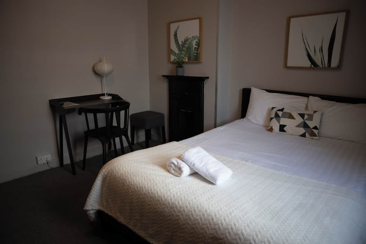 Quarto de hotel com uma cama, uma escrivaninha, uma cadeira, um vaso de plantas e obras de arte nas paredes.