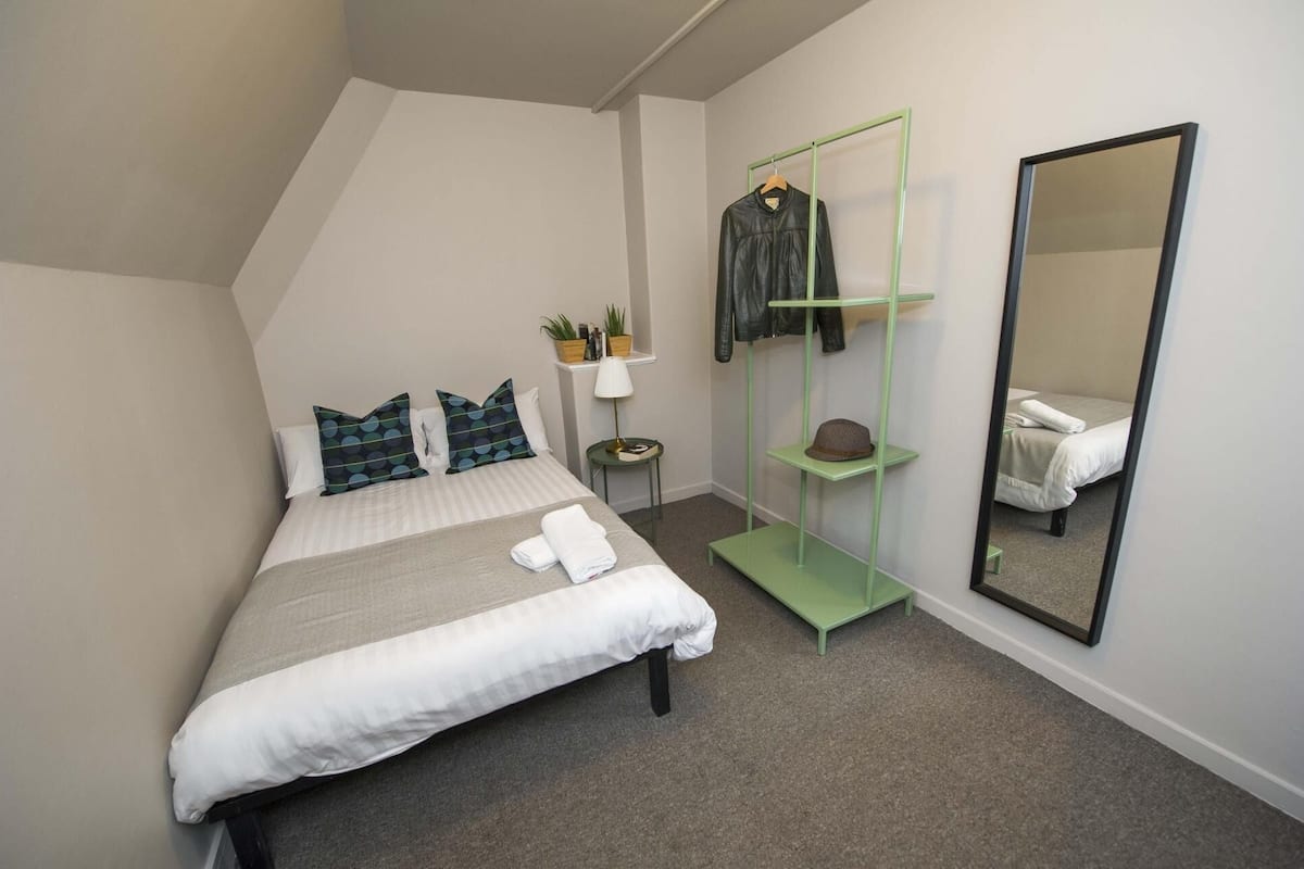 Quarto com cama, almofadas, prateleira verde com espelho e uma pequena mesa com luminária.