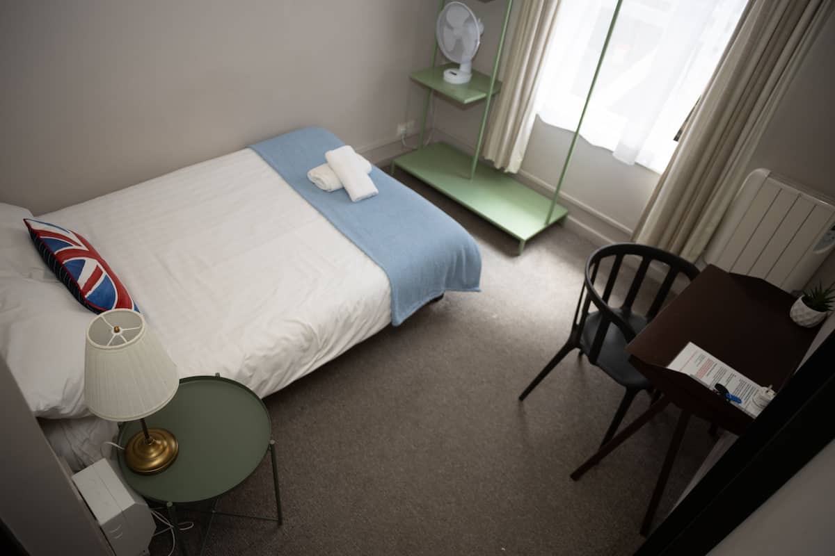 Quarto de hotel com cama, escrivaninha, cadeira, abajur, mesa pequena, ventilador e janela com cortinas.