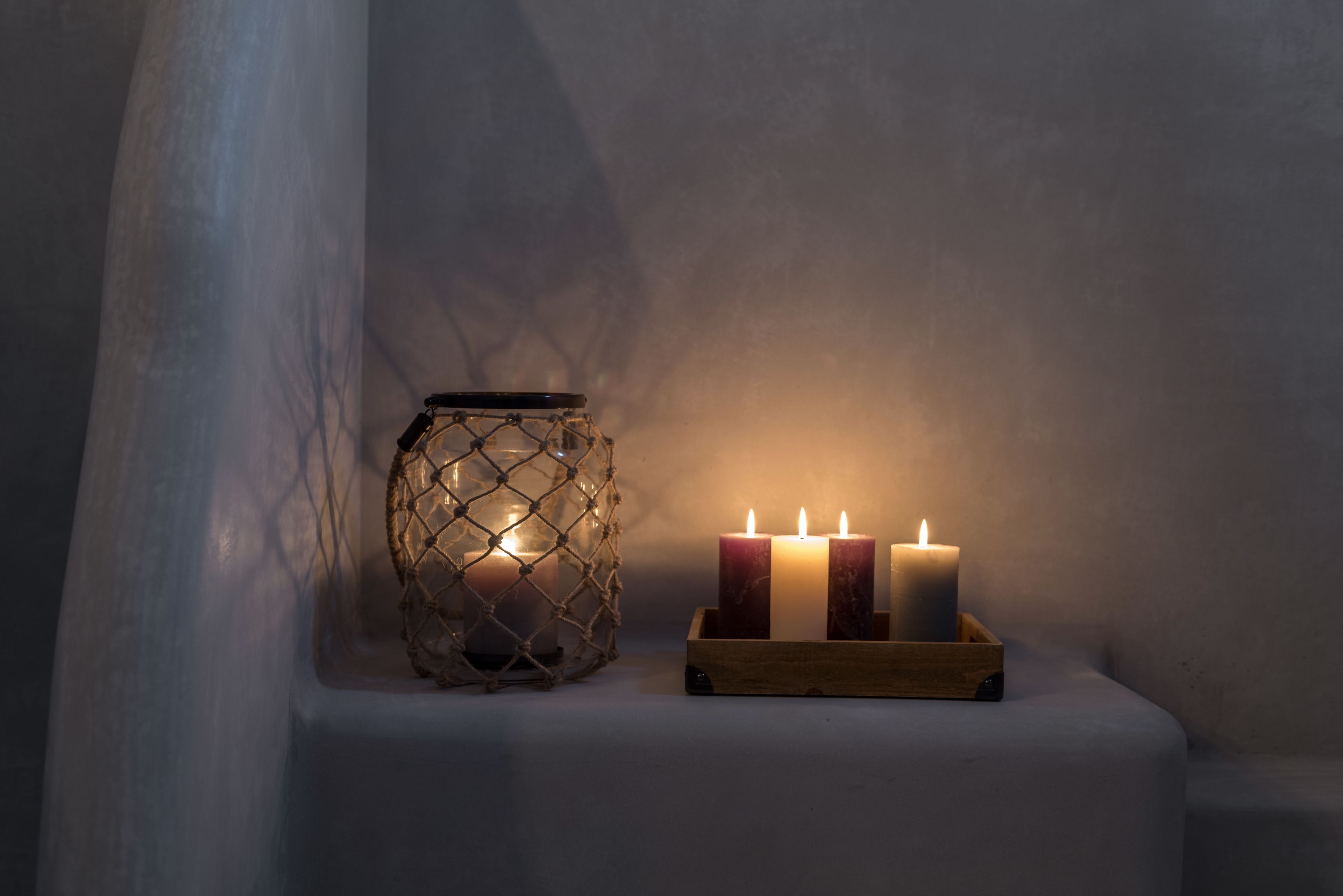 Um canto iluminado por velas, com uma bandeja de madeira que contém três velas e um suporte de vela de rede.