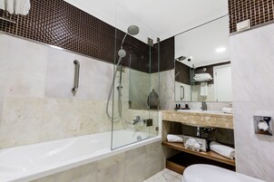 Um banheiro moderno com chuveiro em gabinete de vidro, uma banheira branca, parede revestida de mármore, toucador de madeira com pia e vaso sanitário.