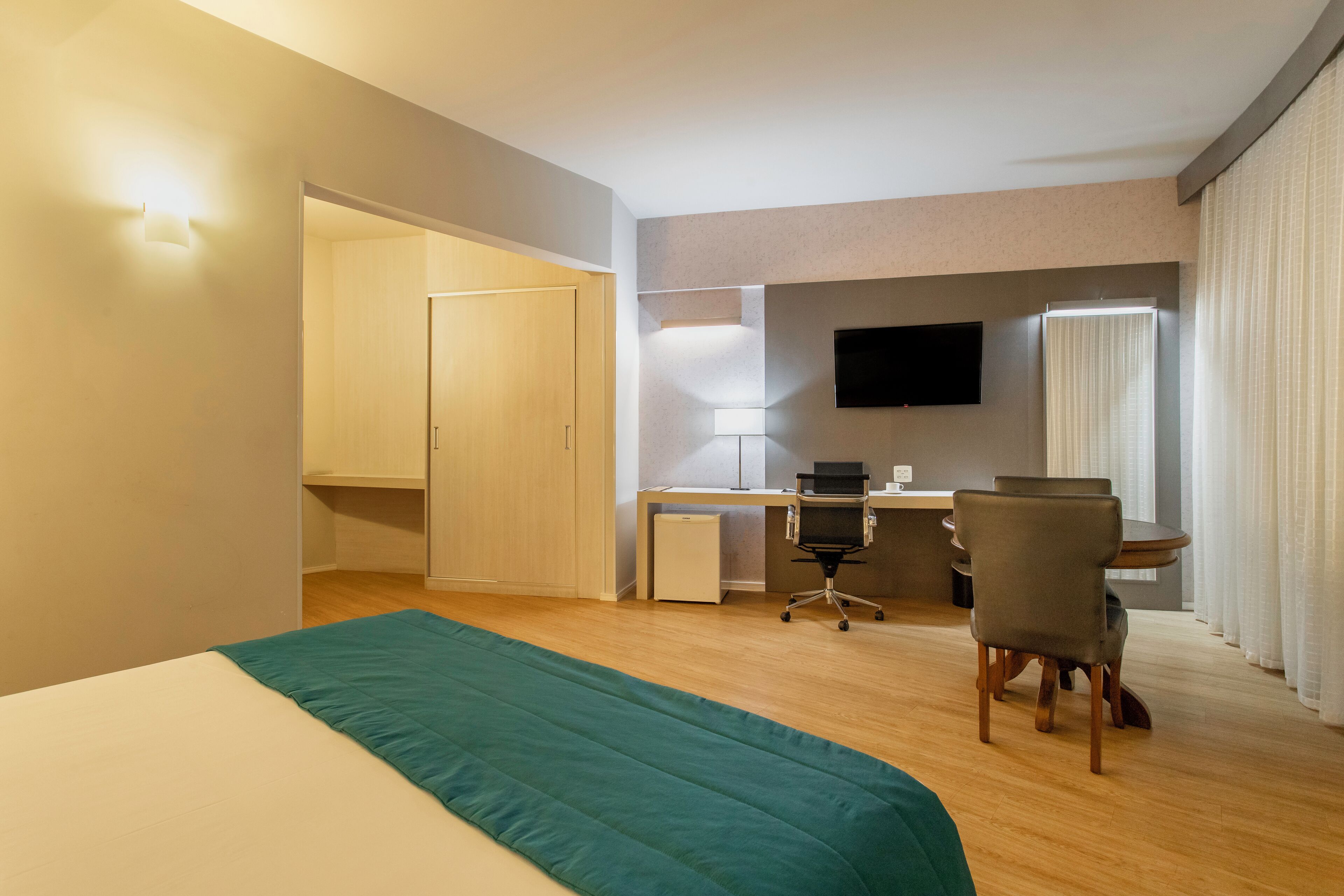 Quarto de hotel com cama, escrivaninha, cadeira, televisão e porta que dá para outro cômodo.