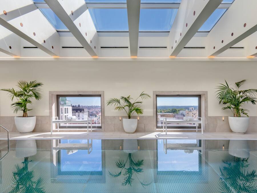 Piscina interior moderna com paredes de vidro transparente, plantas em vasos e vista para a cidade.