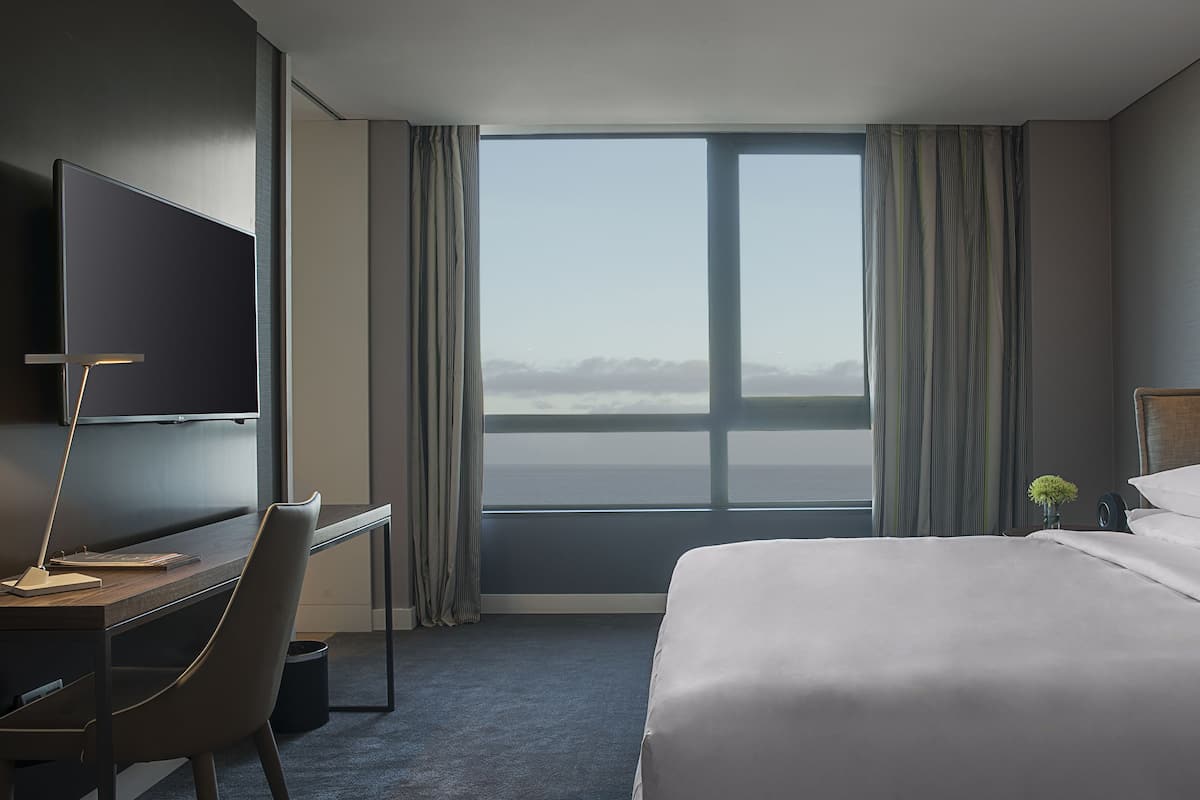 Quarto de hotel com cama, secretária com cadeira, televisão e uma janela grande com cortinas.