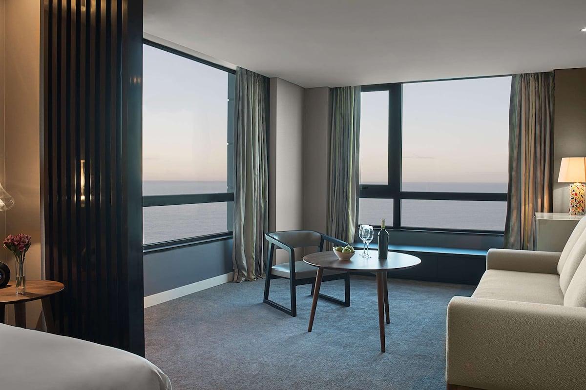 Quarto de hotel com uma grande janela dando vista para o mar, uma mesa redonda com uma cadeira, um sofá e um abajur sobre uma mesinha lateral.