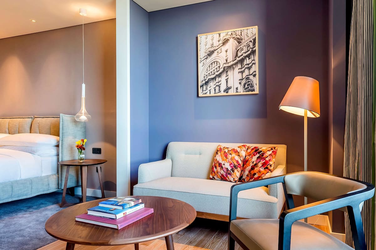 Quarto de hotel moderno com sofá, mesa de centro com livros, luminária e um quadro na parede.