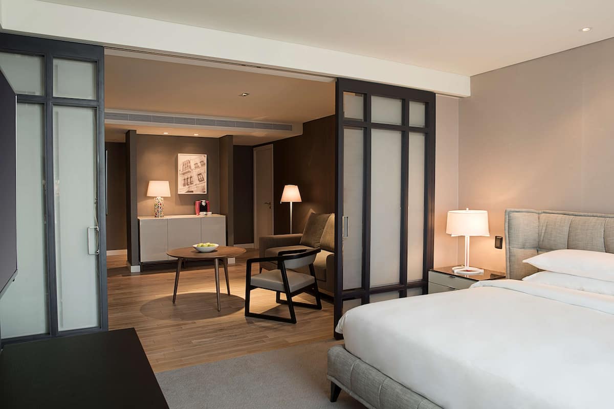 Quarto de hotel com cama, escrivaninha, cadeira, mesa de cabeceira e pequena mesa com abajur.