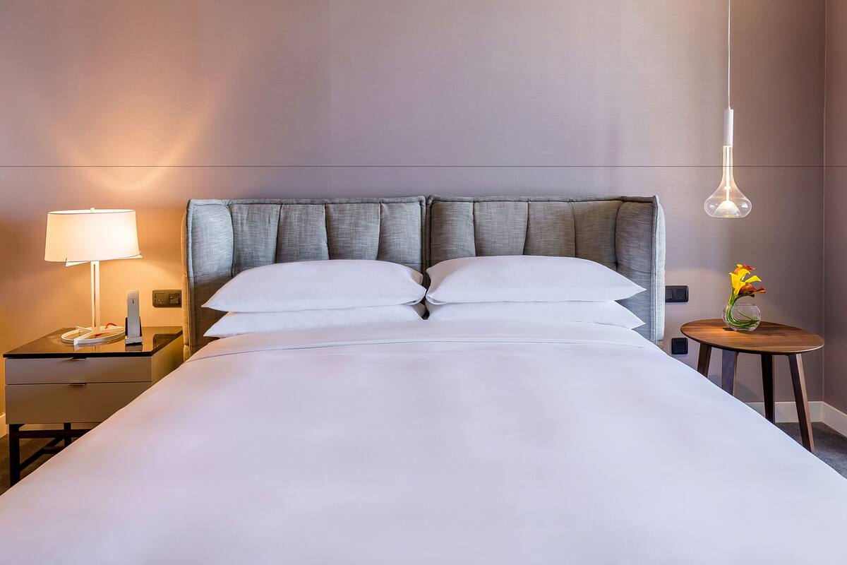 Uma cama bem arrumada com roupa de cama branca, uma mesa de cabeceira com abajur e uma pequena mesa com um vaso de flores.
