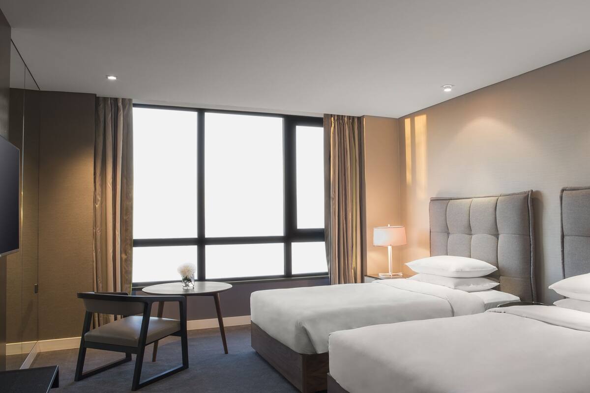Quarto de hotel com duas camas, uma mesinha, uma cadeira, televisão e uma janela grande com cortinas.