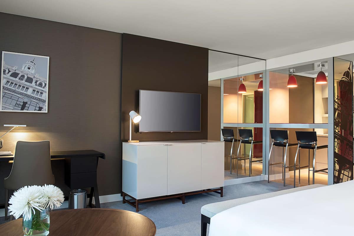 Quarto de hotel moderno com uma cama, uma escrivaninha com luminária, uma cadeira, uma TV e um painel de vidro que dá para uma área de jantar com cadeiras e uma luminária pendente vermelha.