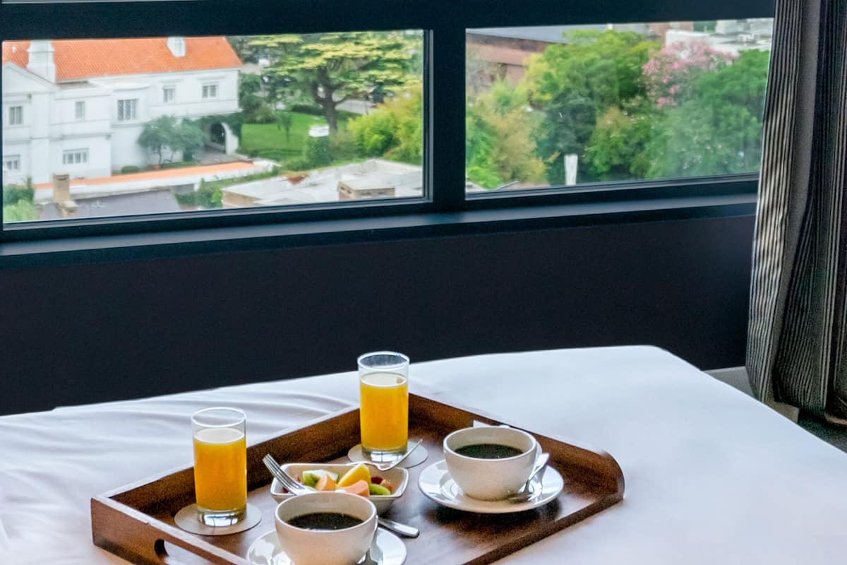 Bandeja de café da manhã com café e suco sobre uma cama de hotel com vista para a cidade.
