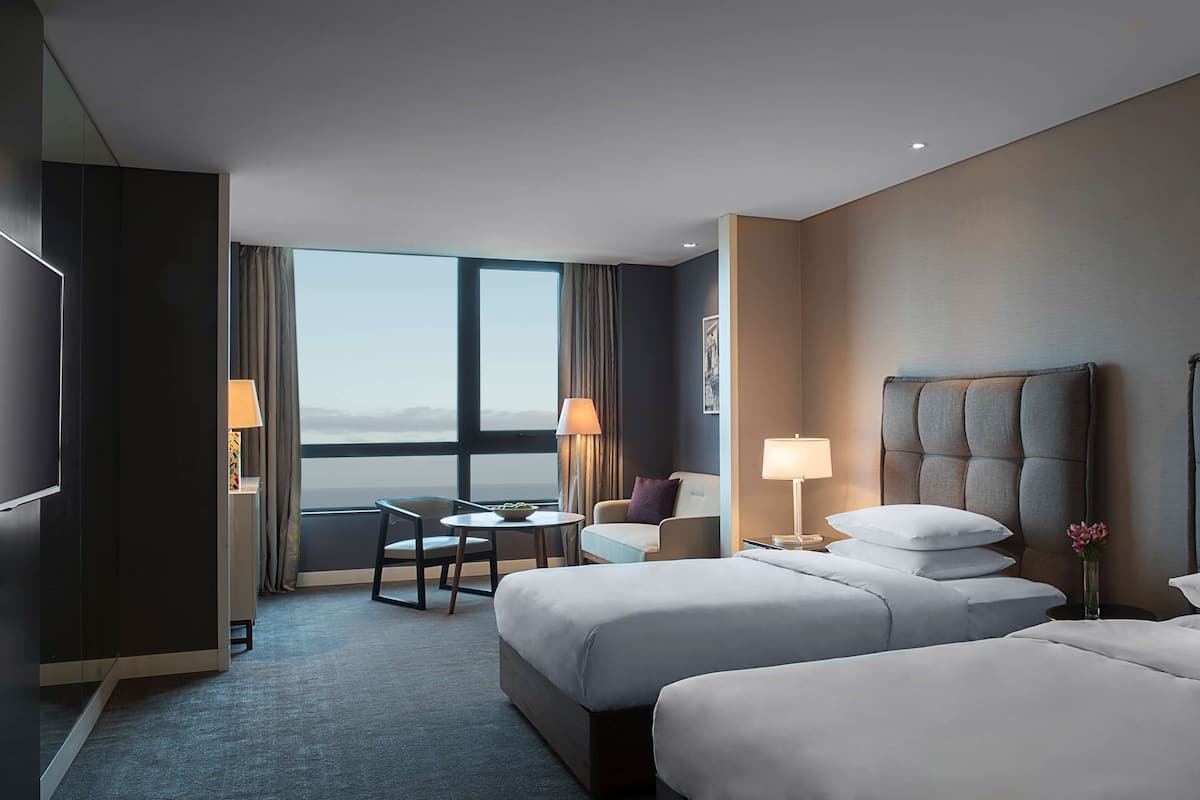 Quarto de hotel com duas camas, uma escrivaninha, uma cadeira e vista para a cidade.