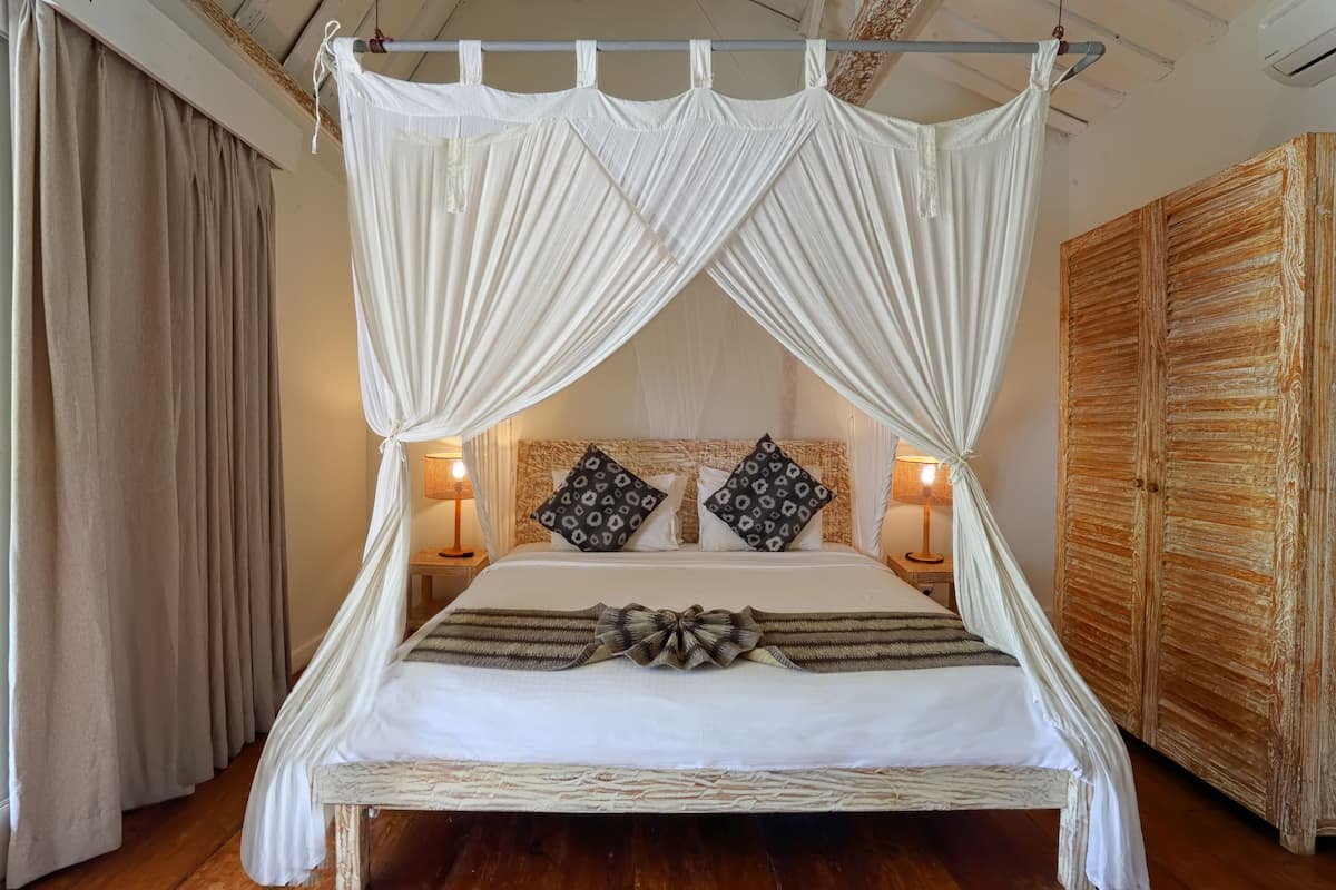 Uma cama de dossel com um edredom branco e um laço decorativo, com duas luminárias de cabeceira e almofadas estampadas de cada lado.