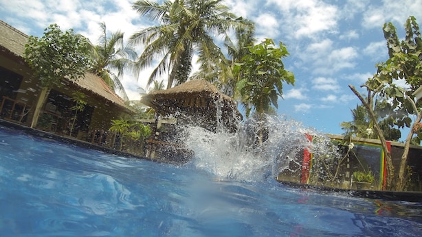 Piscina com água espirrando, cercada por palmeiras e estruturas com telhado de palha.