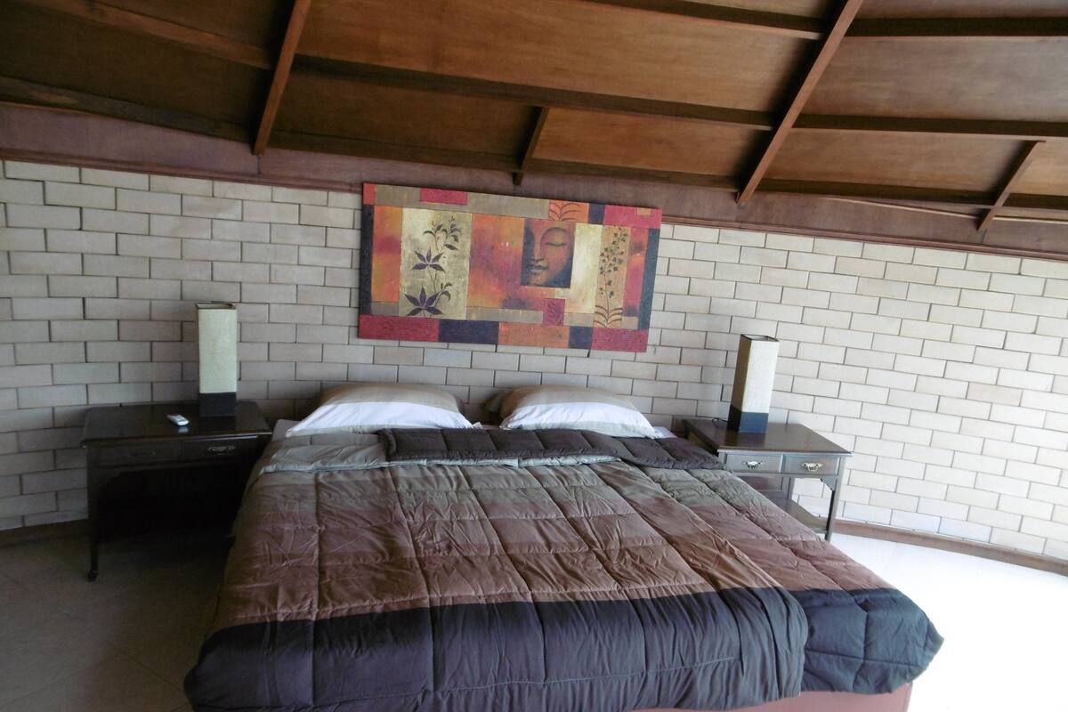 Quarto com cama, duas mesas de cabeceira, uma obra de arte na parede e parede de tijolos.