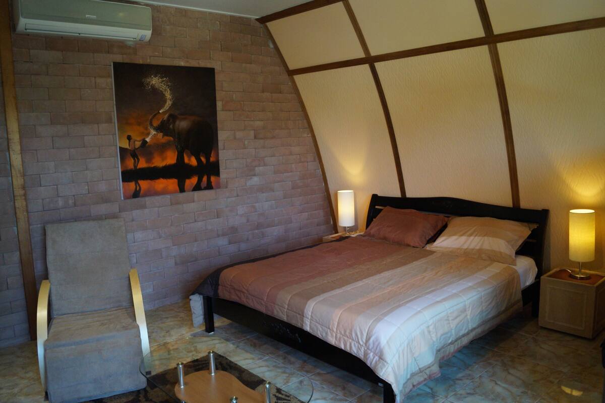 Quarto com cama, poltrona, mesa de cabeceira, luminária e um quadro na parede.