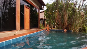 Piscina com borda azulejada, cercada por vegetação e um edifício com porta de madeira e janelas amplas.