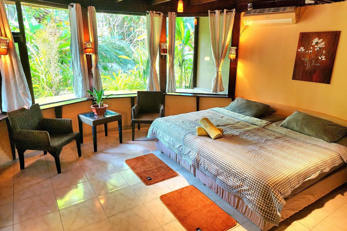 Quarto com cama, duas cadeiras, uma mesinha com planta em vaso e janela com cortinas.