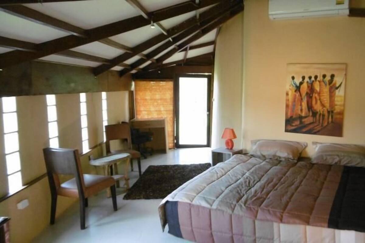 Quarto com cama, escrivaninha e cadeira. Há um quadro na parede e uma janela com persianas.