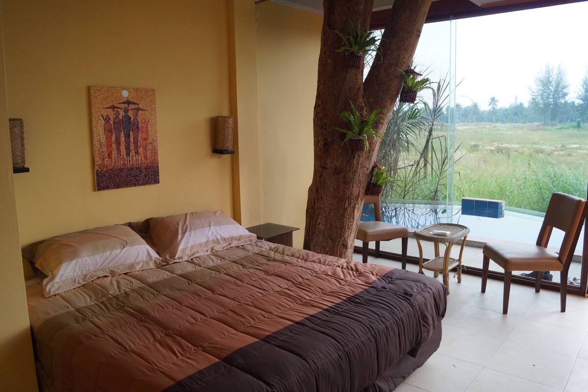 Quarto com uma cama grande, mesa de cabeceira, uma cadeira e uma mesa. Há uma árvore no quarto e vista para o verde lá fora.