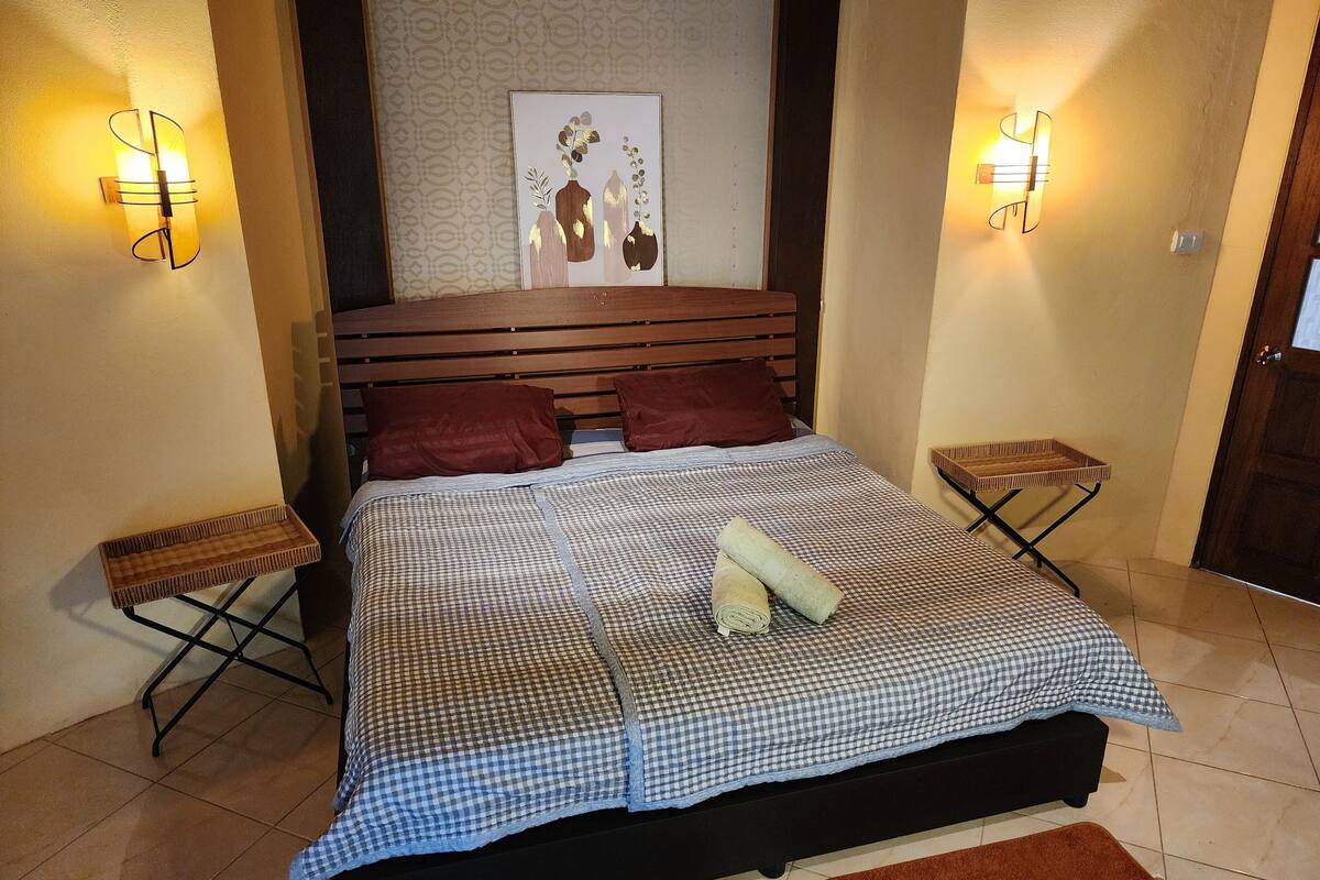 Cama bem arrumada com colcha xadrez, duas almofadas e toalha dobrada. Há duas mesas de cabeceira com abajures, uma cabeceira de madeira e um quadro na parede.