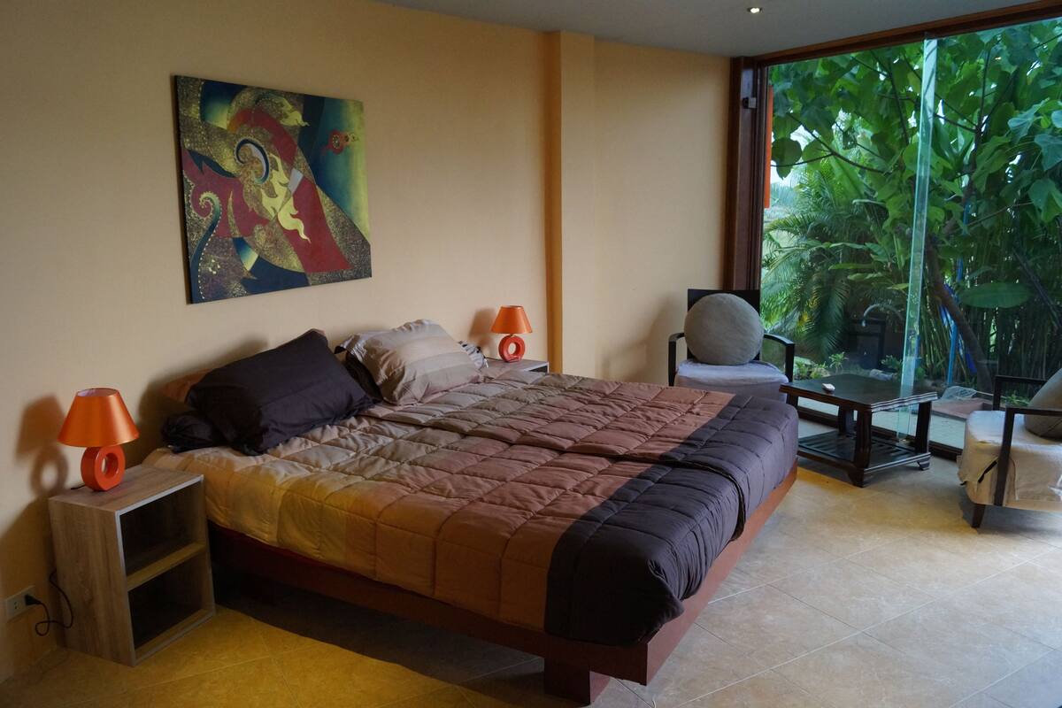 Quarto com uma cama grande, duas mesas de cabeceira com abajures, uma cadeira e uma janela com vista para o verde.