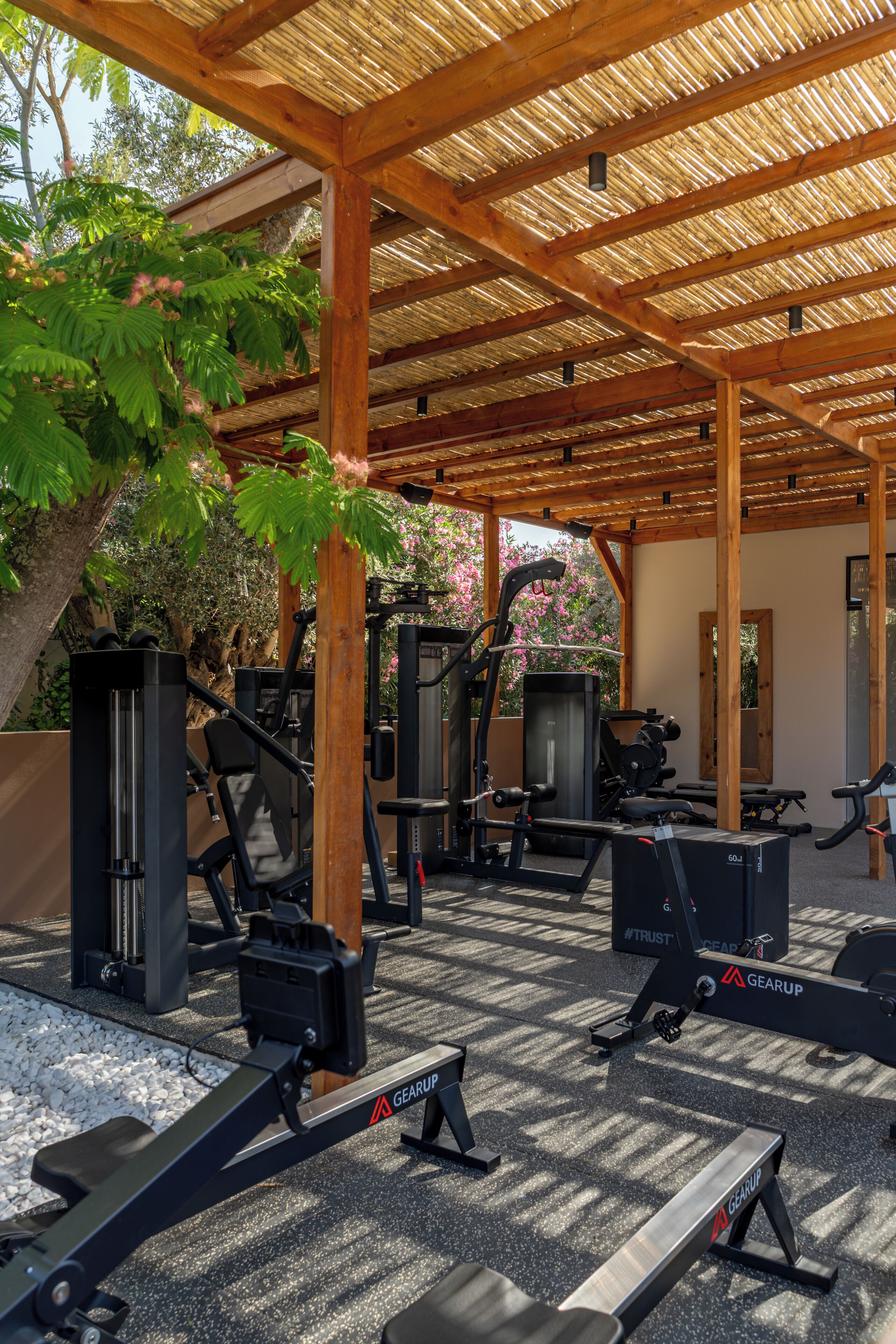 Uma área de ginástica ao ar livre com diversos aparelhos de exercício, uma pérgula de madeira e uma árvore com flores cor-de-rosa.
