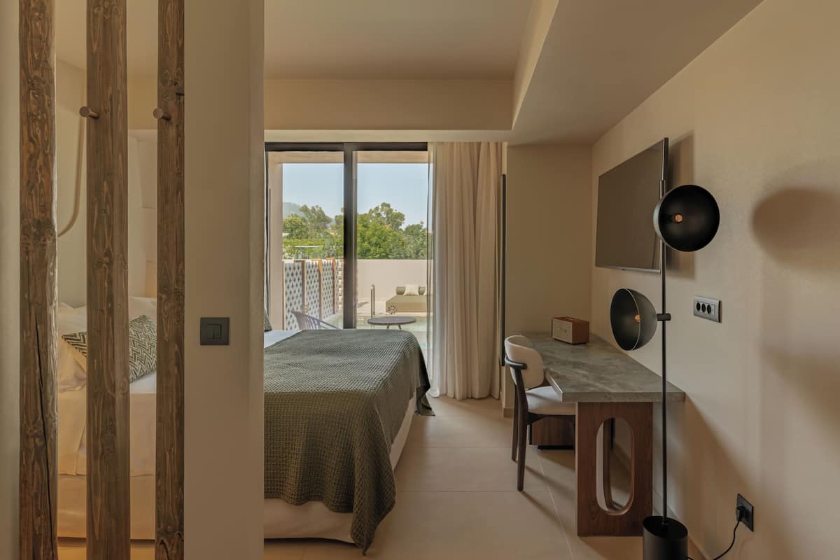 Quarto de hotel com cama, escrivaninha com cadeira, televisão e varanda com mesa e cadeiras.