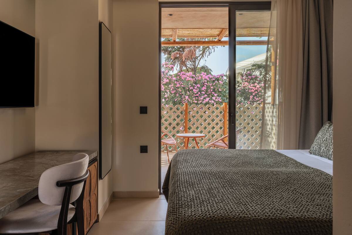Quarto com cama, escrivaninha e cadeira. Possui varanda com vista para flores cor-de-rosa e pérgola de madeira.