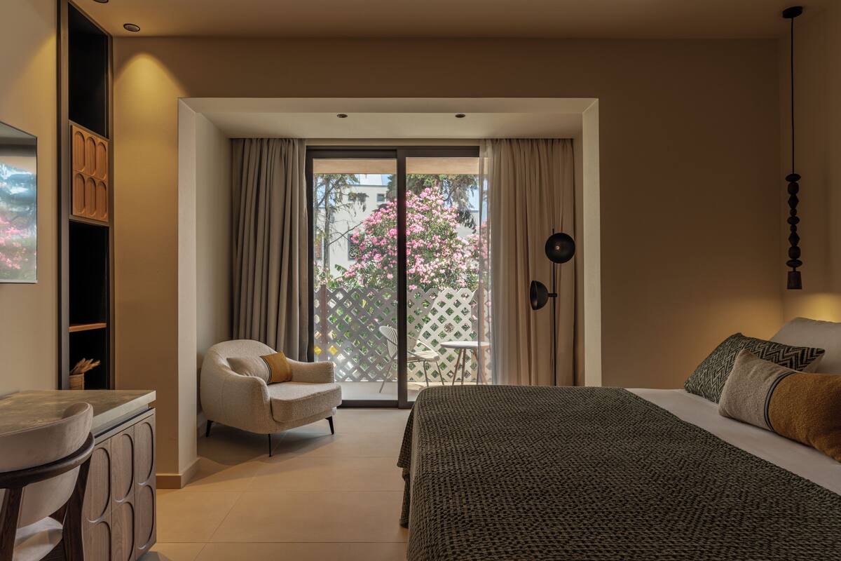Quarto com cama, uma cadeira, uma escrivaninha e vista para uma varanda com flores cor-de-rosa.