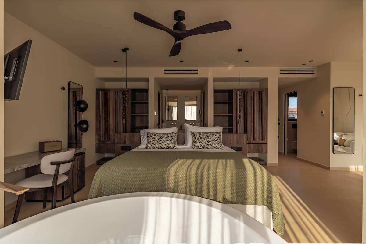 Quarto espaçoso com uma cama grande, uma escrivaninha e uma cadeira. Há um ventilador de teto, um espelho e um guarda-roupa.
