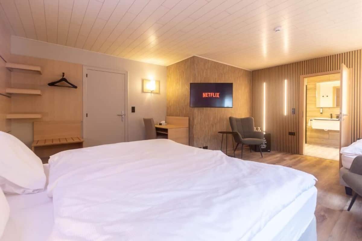 Quarto de hotel com cama, escrivaninha, cadeira, TV fixada na parede e painel de parede de madeira.