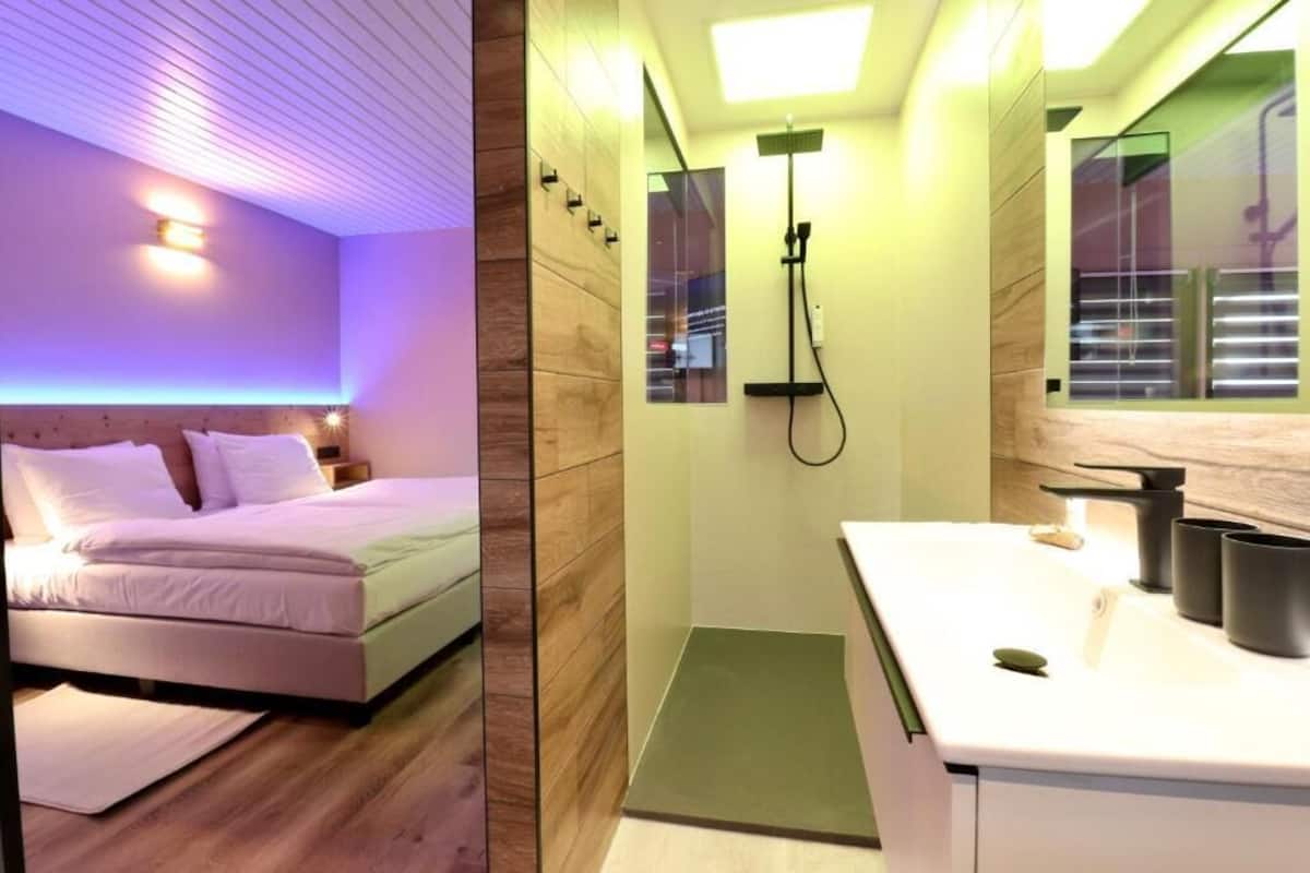Quarto moderno com cama, mesa de cabeceira e luminária na parede. A imagem também mostra um banheiro com chuveiro, pia e espelho.