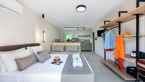Quarto de hotel moderno com uma cama, mesa de cabeceira com toalhas, prateleira com roupas e vista para a cozinha.