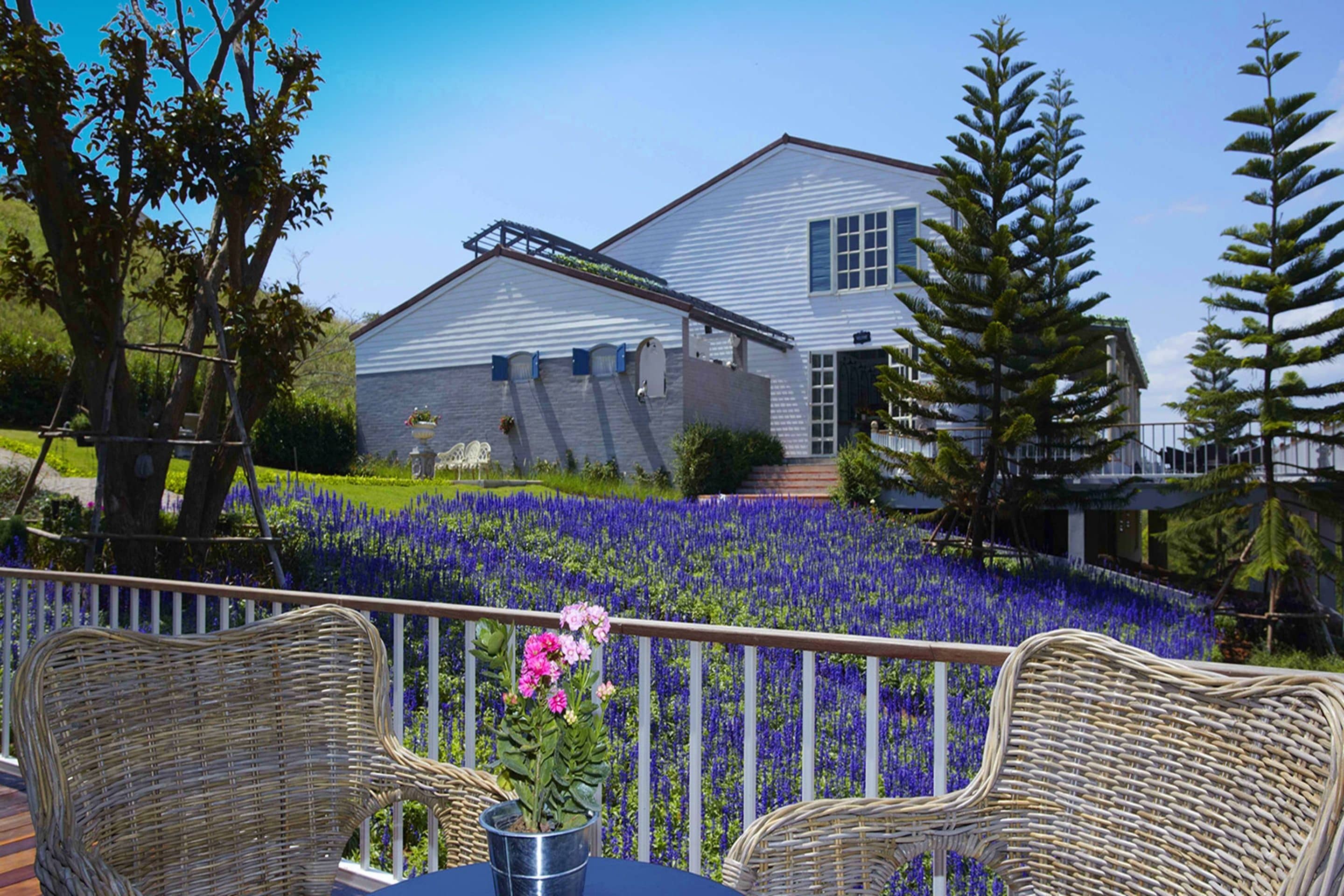 Uma casa com varanda e vista para um jardim com flores roxas.