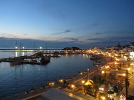 Uma marina costeira com barcos atracados, uma ponte ao fundo e uma paisagem urbana com edifícios e iluminação pública.
