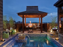 Um cozinheiro preparando refeições ao ar livre num gazebo de madeira, com uma piscina em primeiro plano.