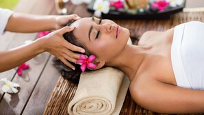 Uma pessoa recebendo massagem na cabeça em um spa, com uma mesa de madeira, toalha enrolada e flores.