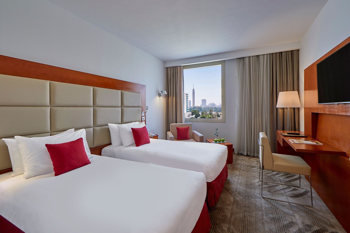 Quarto de hotel com duas camas, uma escrivaninha, uma cadeira e uma janela grande com vista para a cidade.