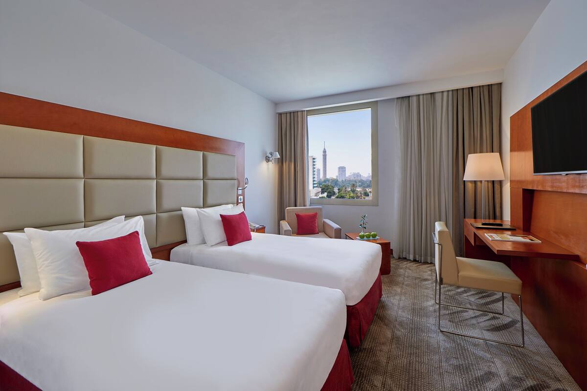 Quarto de hotel com duas camas, uma escrivaninha, uma cadeira, uma televisão e uma janela com vista para a cidade.
