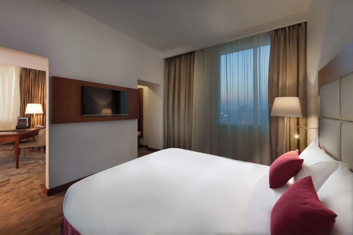 Quarto de hotel com uma cama, televisão, escrivaninha e vista para a cidade ao entardecer.
