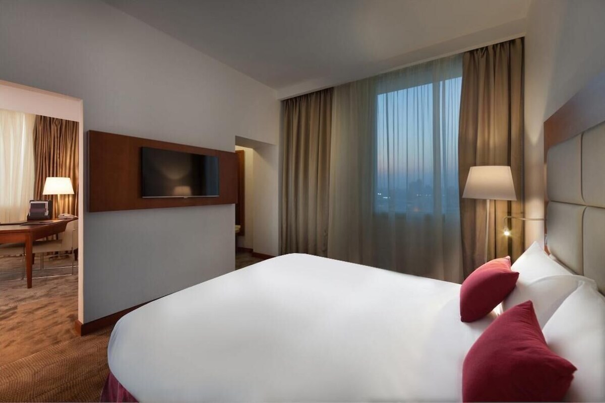 Quarto de hotel com uma cama grande, uma escrivaninha com luminária, uma televisão e vista para a cidade pela janela.