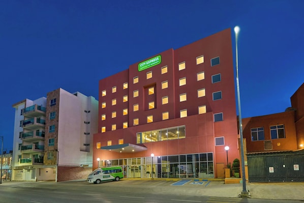 Um hotel com vários andares, com um grande letreiro da City Express, janelas amplas e um estacionamento com um ônibus verde.
