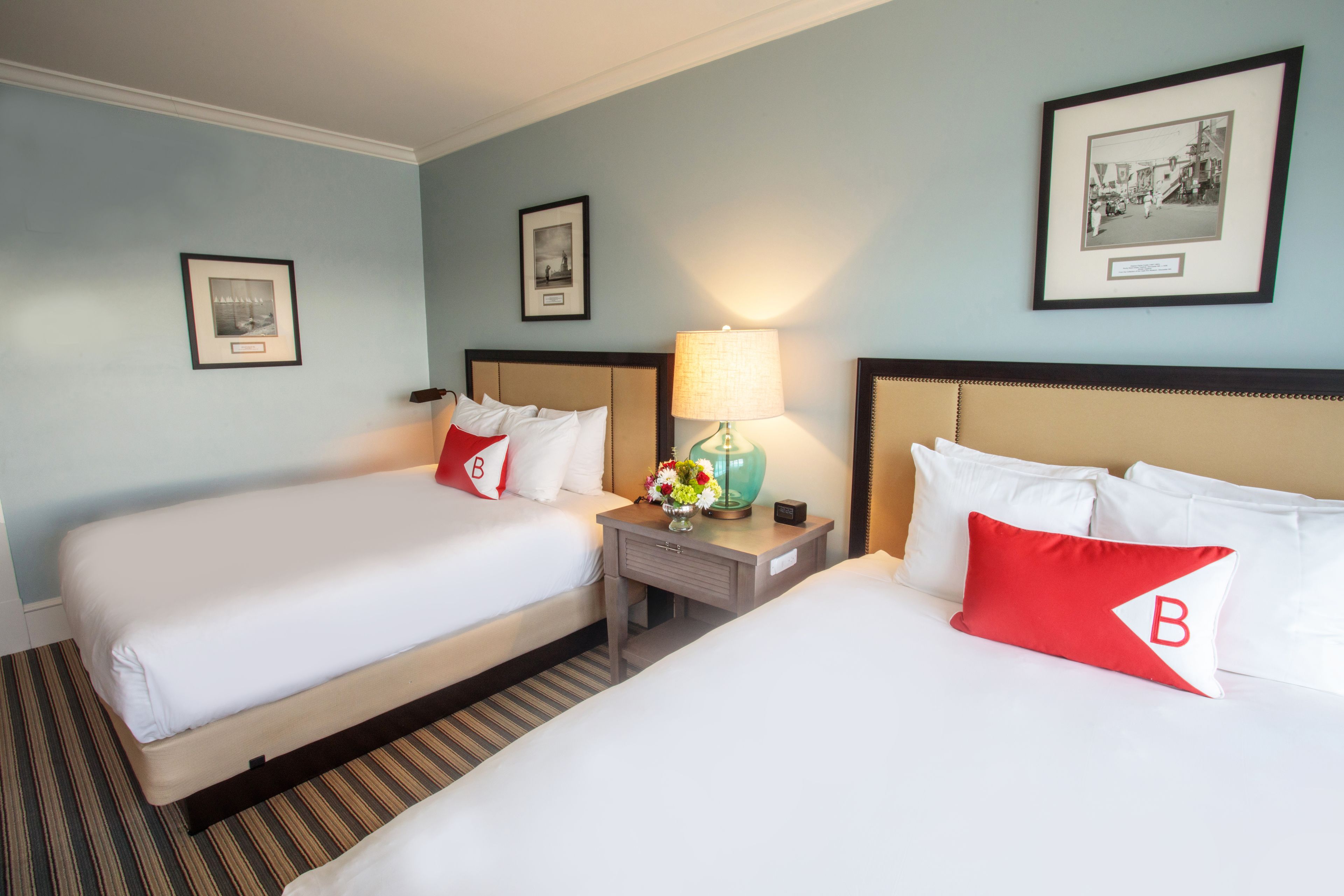 Quarto de hotel com duas camas, mesa de cabeceira com abajur e quadros nas paredes.
