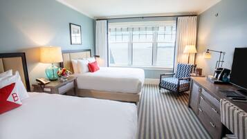 Quarto de hotel com duas camas, uma escrivaninha, uma cadeira, uma televisão e uma janela com vista para o mar.