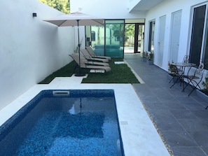 Piscina com espreguiçadeiras e guarda-sol, pequena área ajardinada e porta que dá para um alpendre coberto.