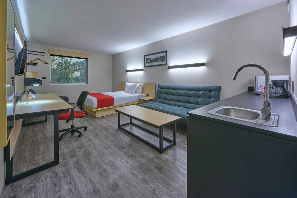 Quarto de hotel moderno com cama, sofá, escrivaninha e cozinha compacta.
