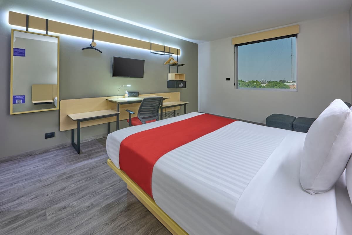 Quarto de hotel com cama, escrivaninha, cadeira e televisão fixada na parede.