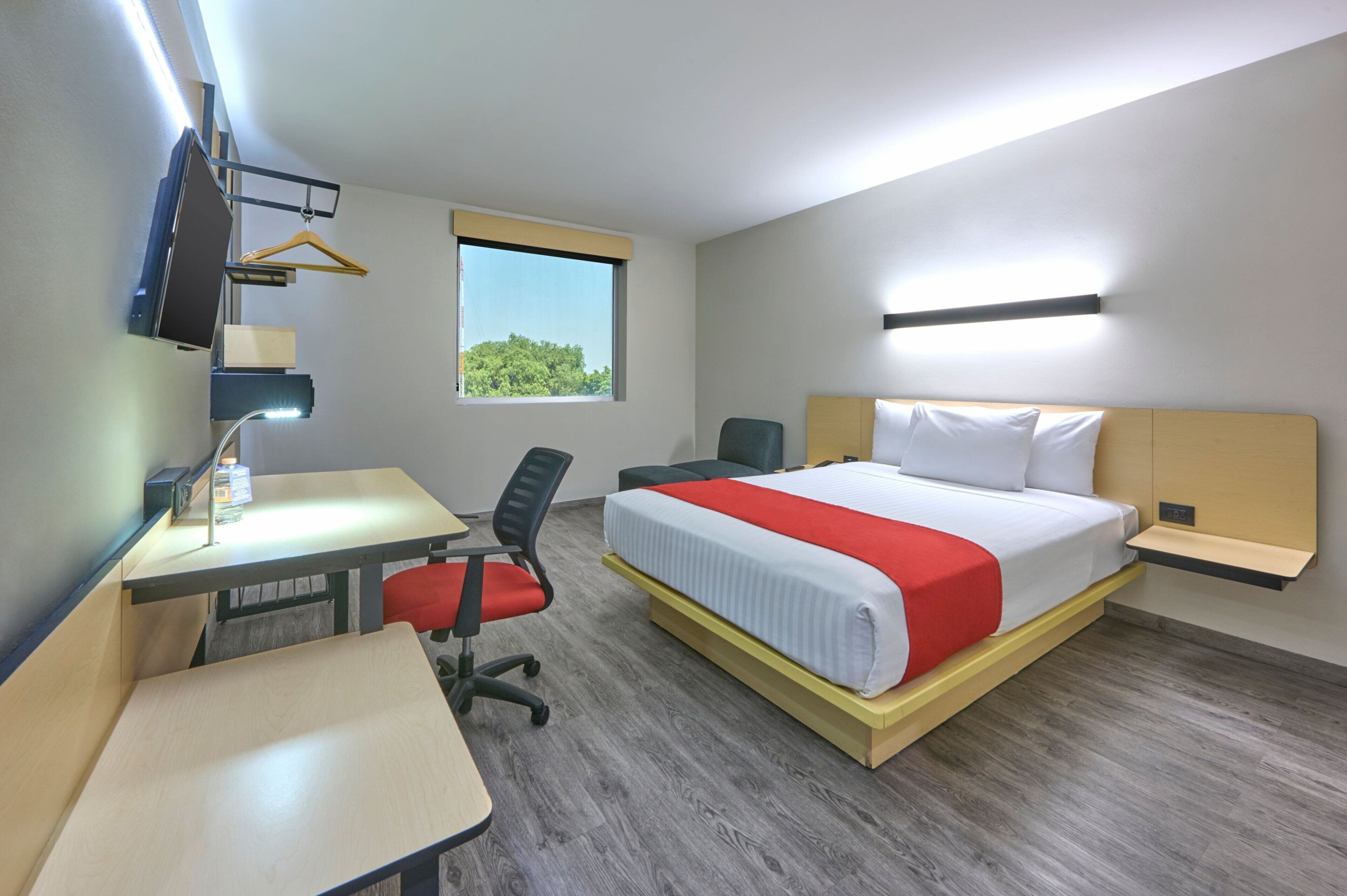 Quarto de hotel com cama, escrivaninha, cadeira e televisão fixada na parede.
