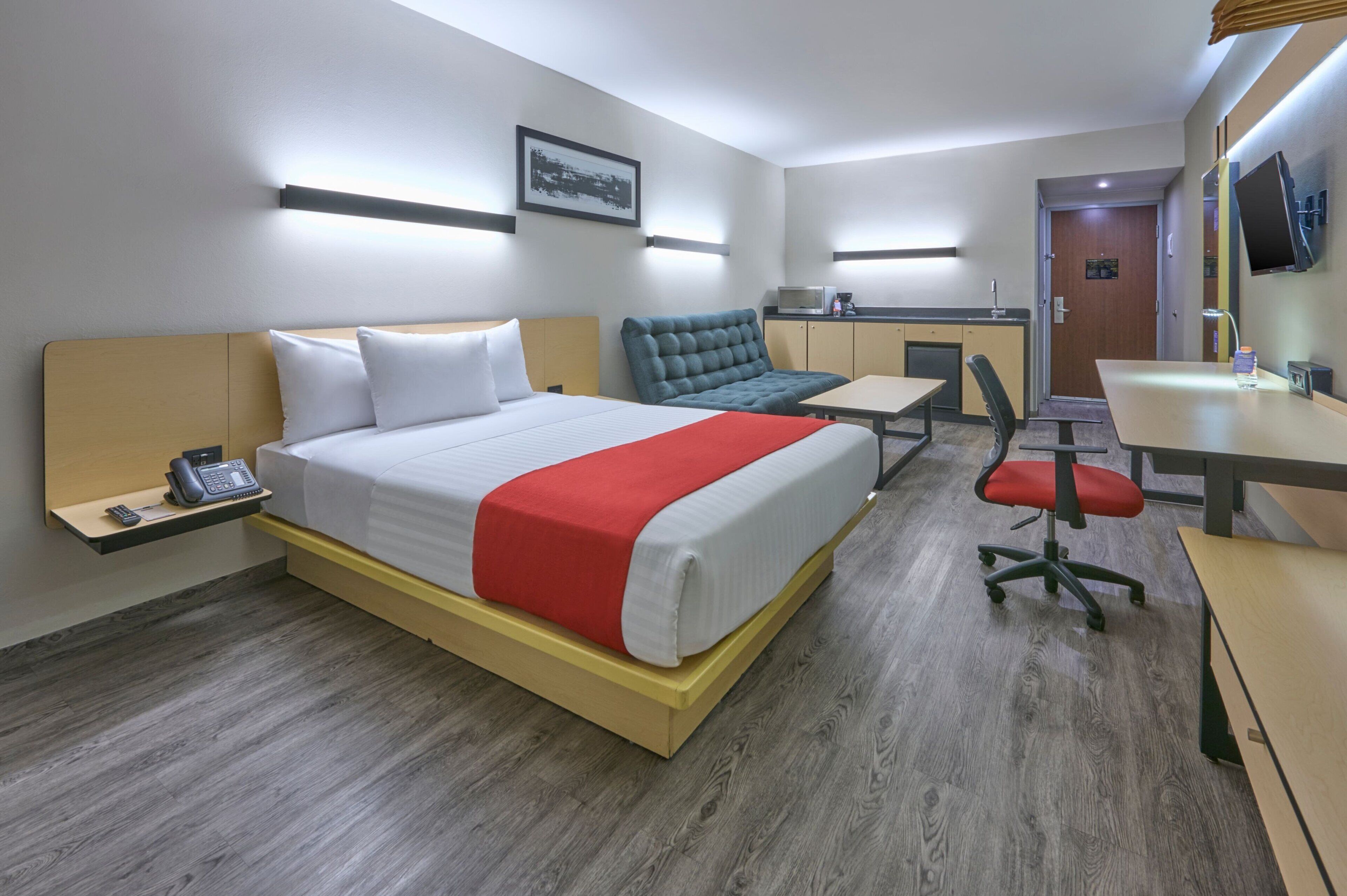 Quarto de hotel com cama, mesa de cabeceira com telefone, escrivaninha com cadeira, micro-ondas e televisão na parede.